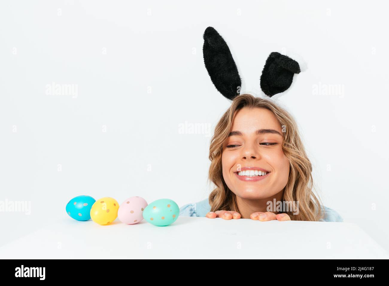 Cheerful young woman wearing easter bunny ears peeking out from table ...