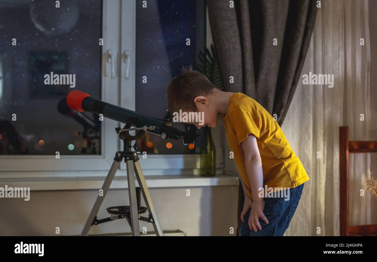 Cute boy is looking through a telescope in a room at the night starry ...