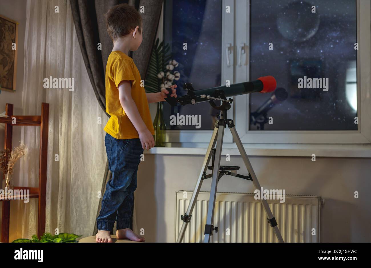Cute boy is looking through a telescope in a room at the night starry ...