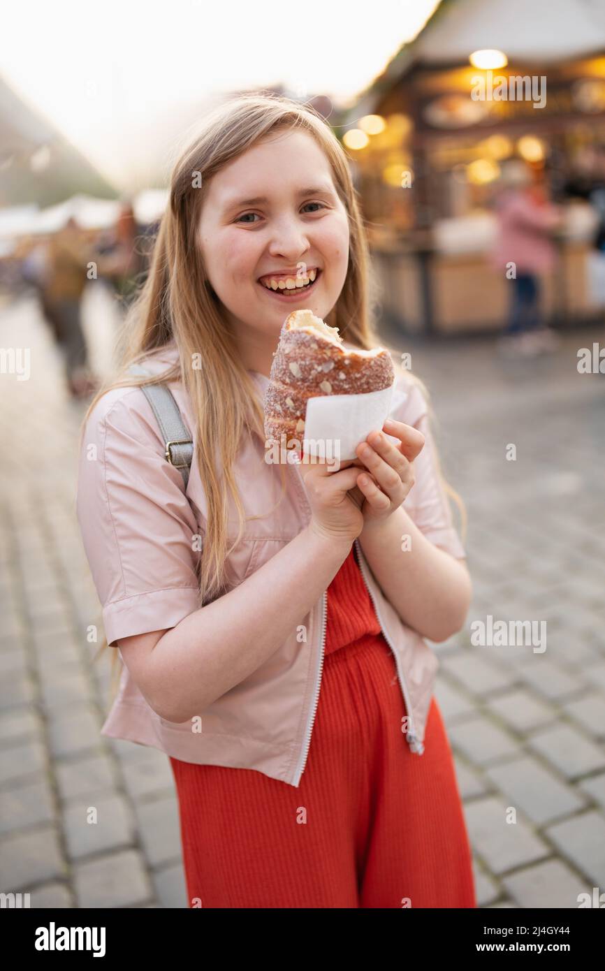 Portrait of smiling modern girl at the fair in the city eating trdelnik ...
