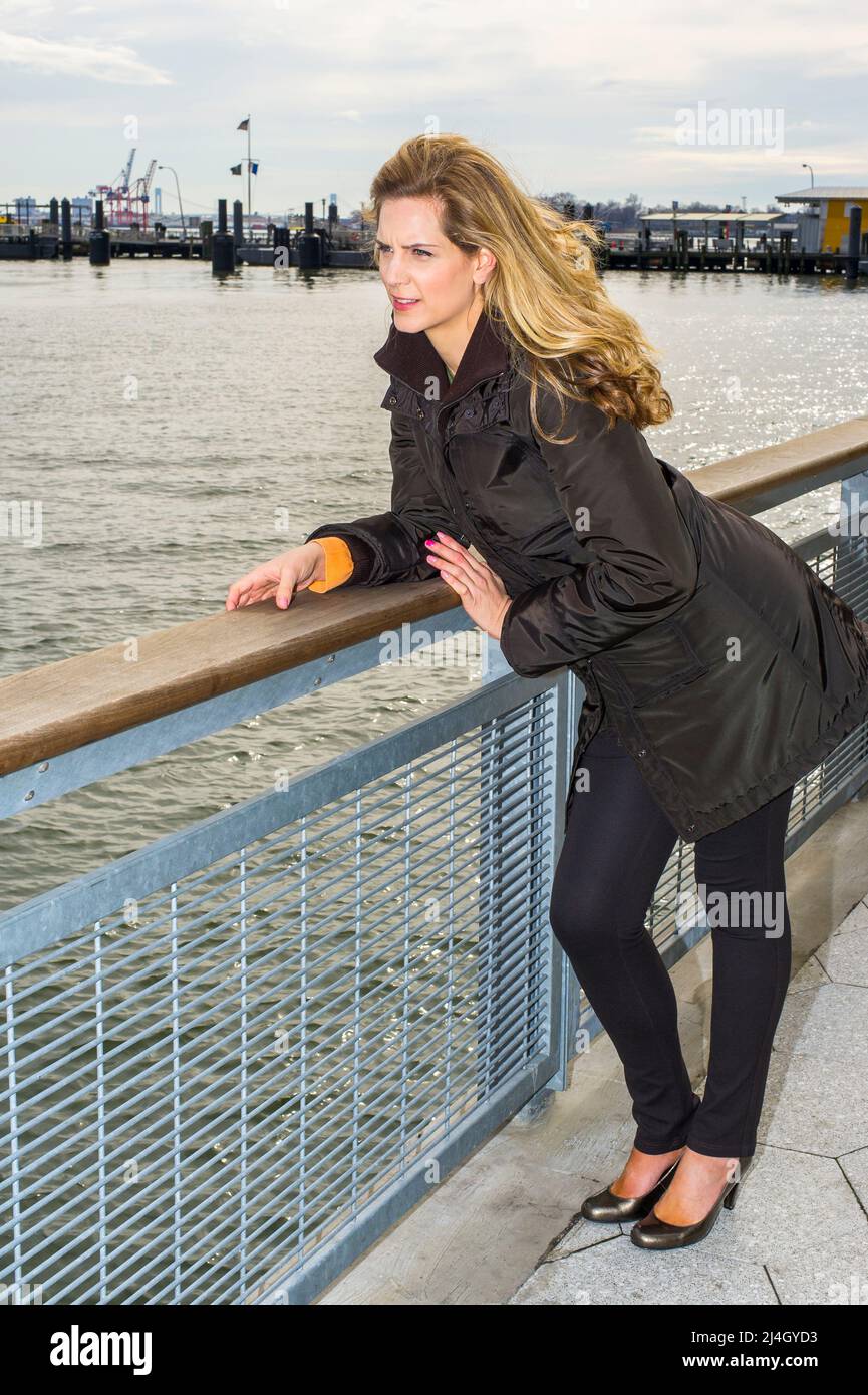 A pretty woman is standing and bending over a fence by a river in a ...