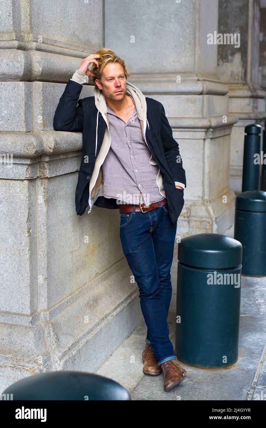 A young handsome guy is standing outside against a column, scratching ...