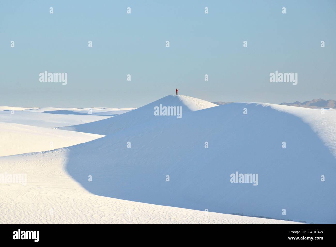 A lone human figure stands atop an immense sand dune under a bright ...