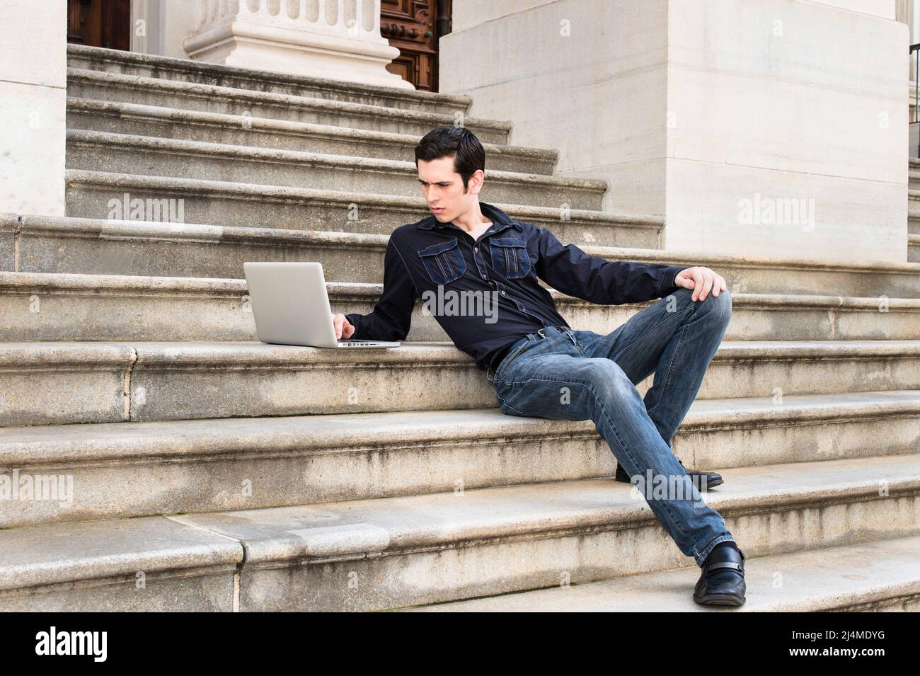 A young handsome guy is sitting on steps and studying on a computer ...