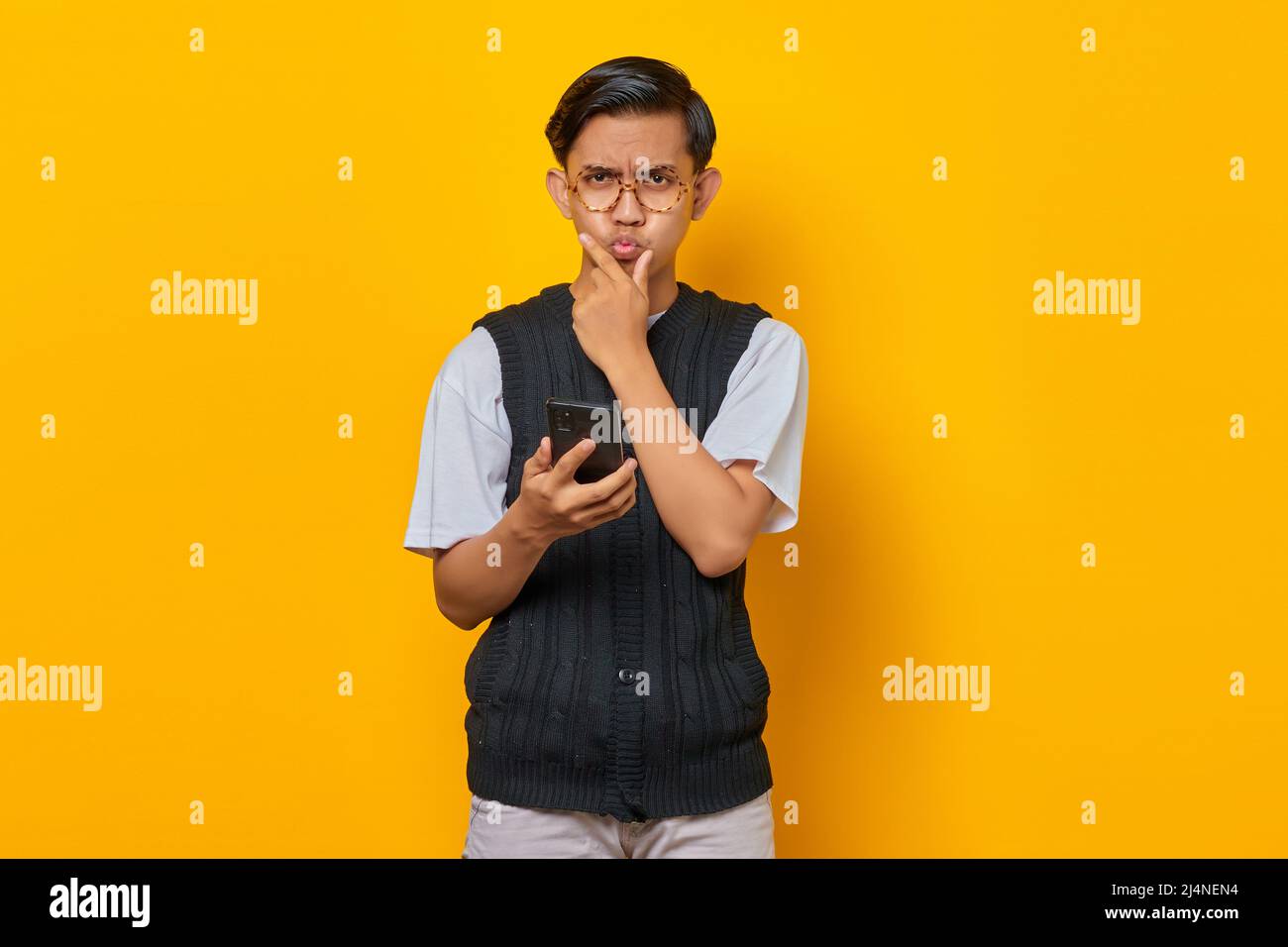 Angry young Asian man with finger on chin and holding mobile phone over ...