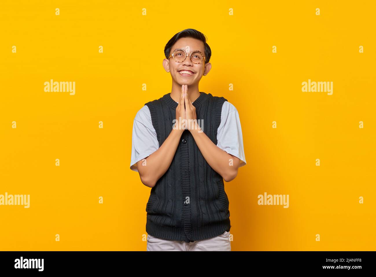 Portrait of smiling Asian young man showing prayer gesture on yellow ...