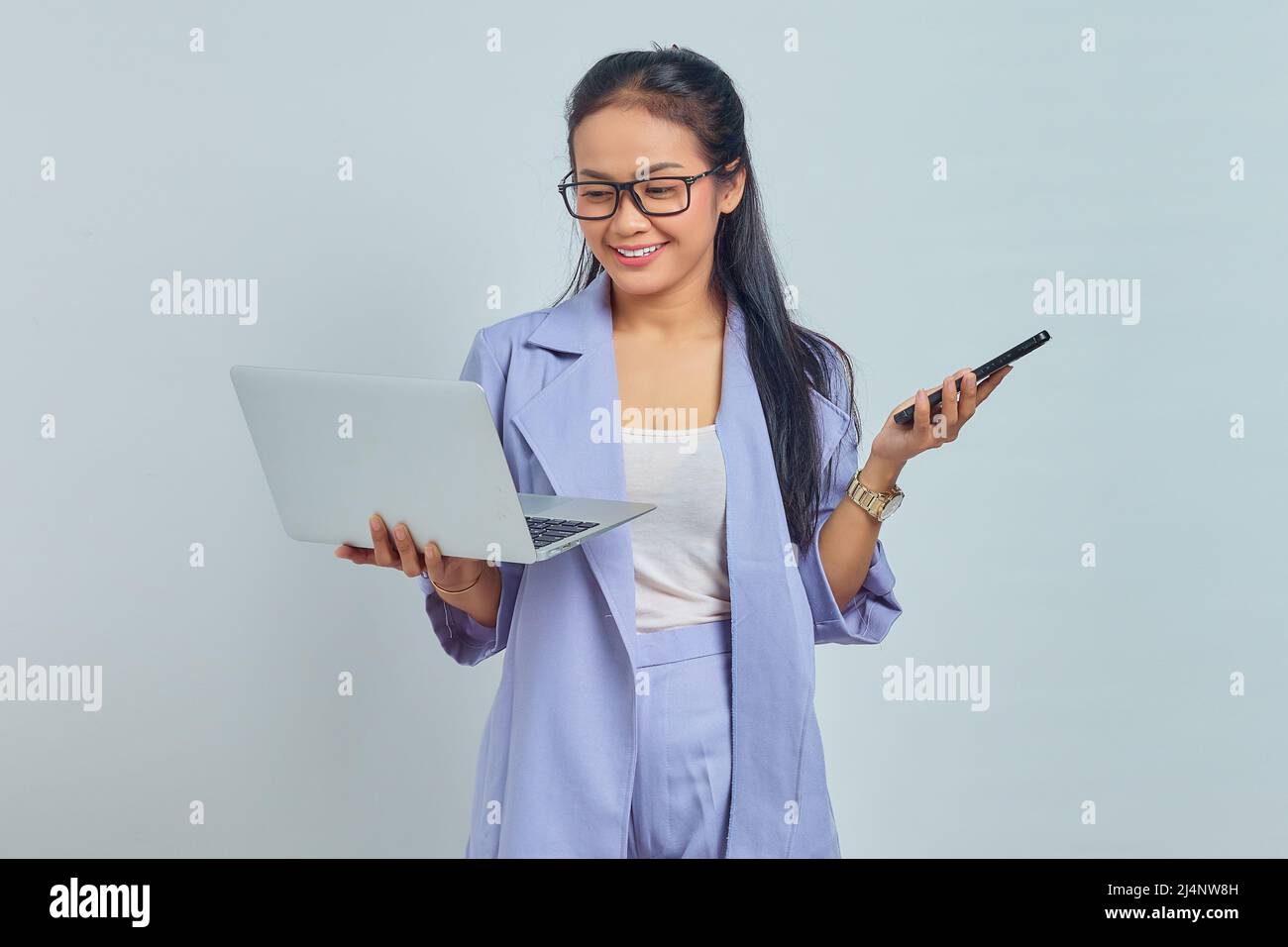 Portrait of young Asian woman standing using laptop and holding mobile ...