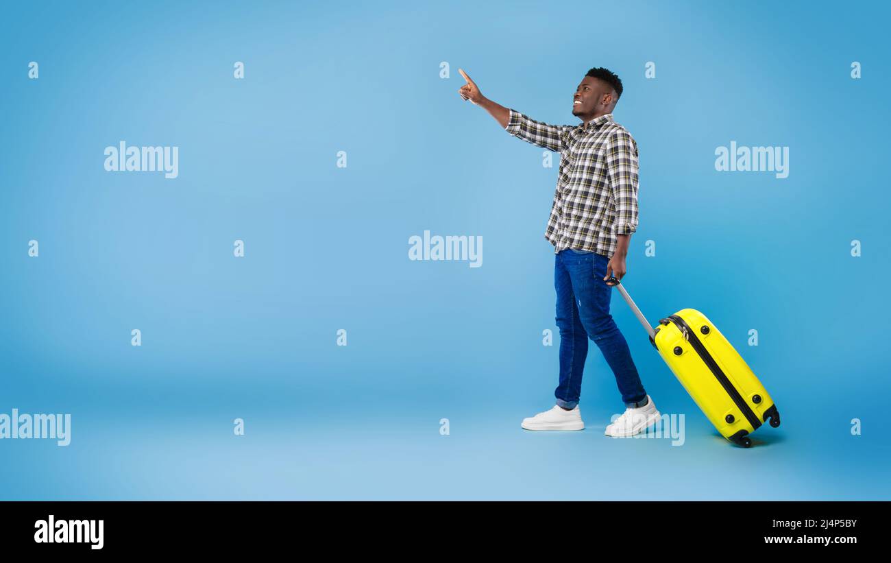 Handsome millennial black guy walking with bright suitcase, pointing at ...