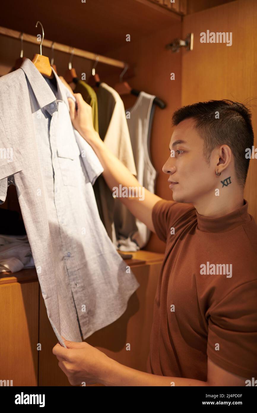 Young man standing in closet and holding hanger with choosing to wear ...