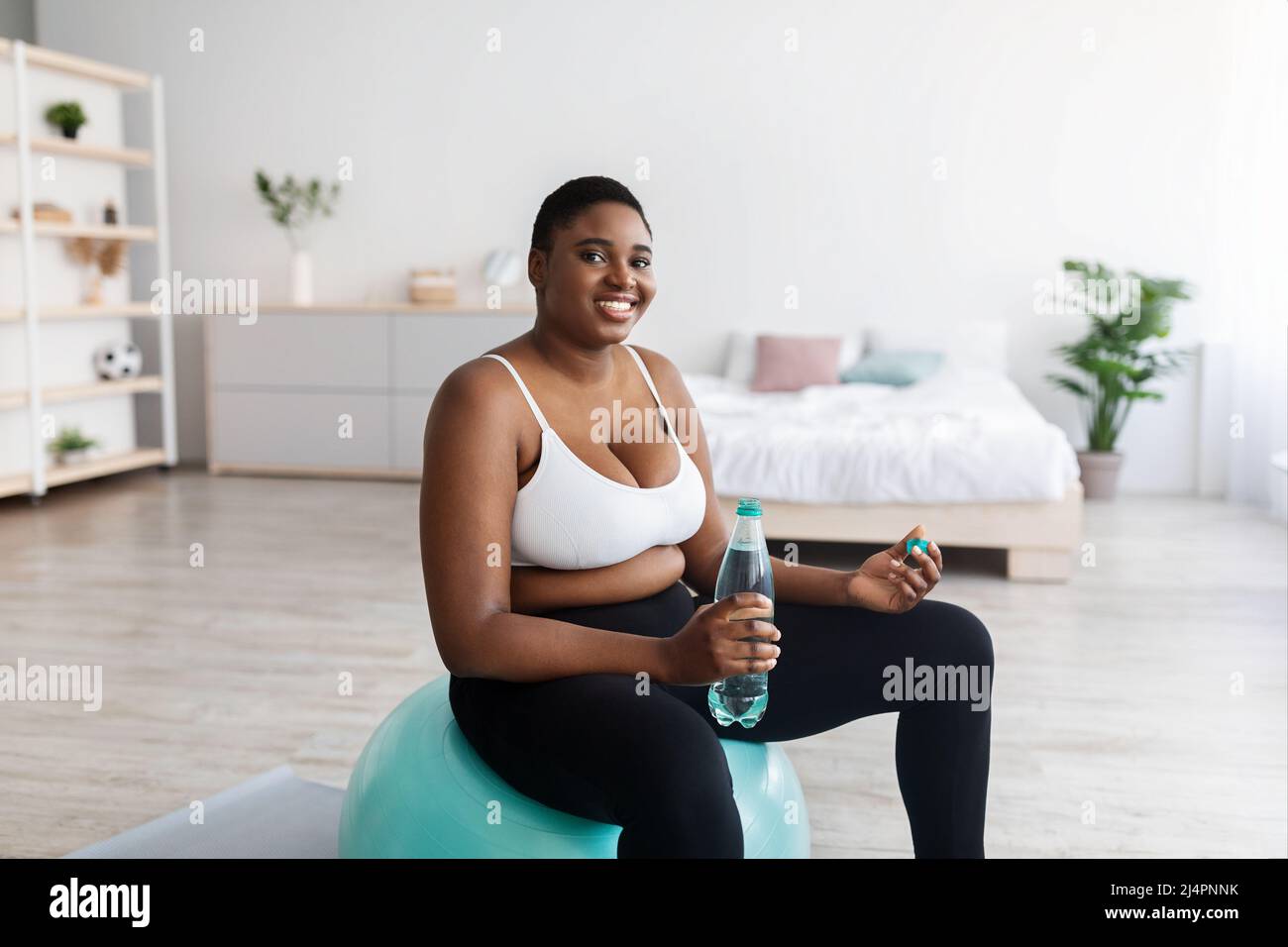 Plus size young black woman sitting in fitness ball with bottle of ...