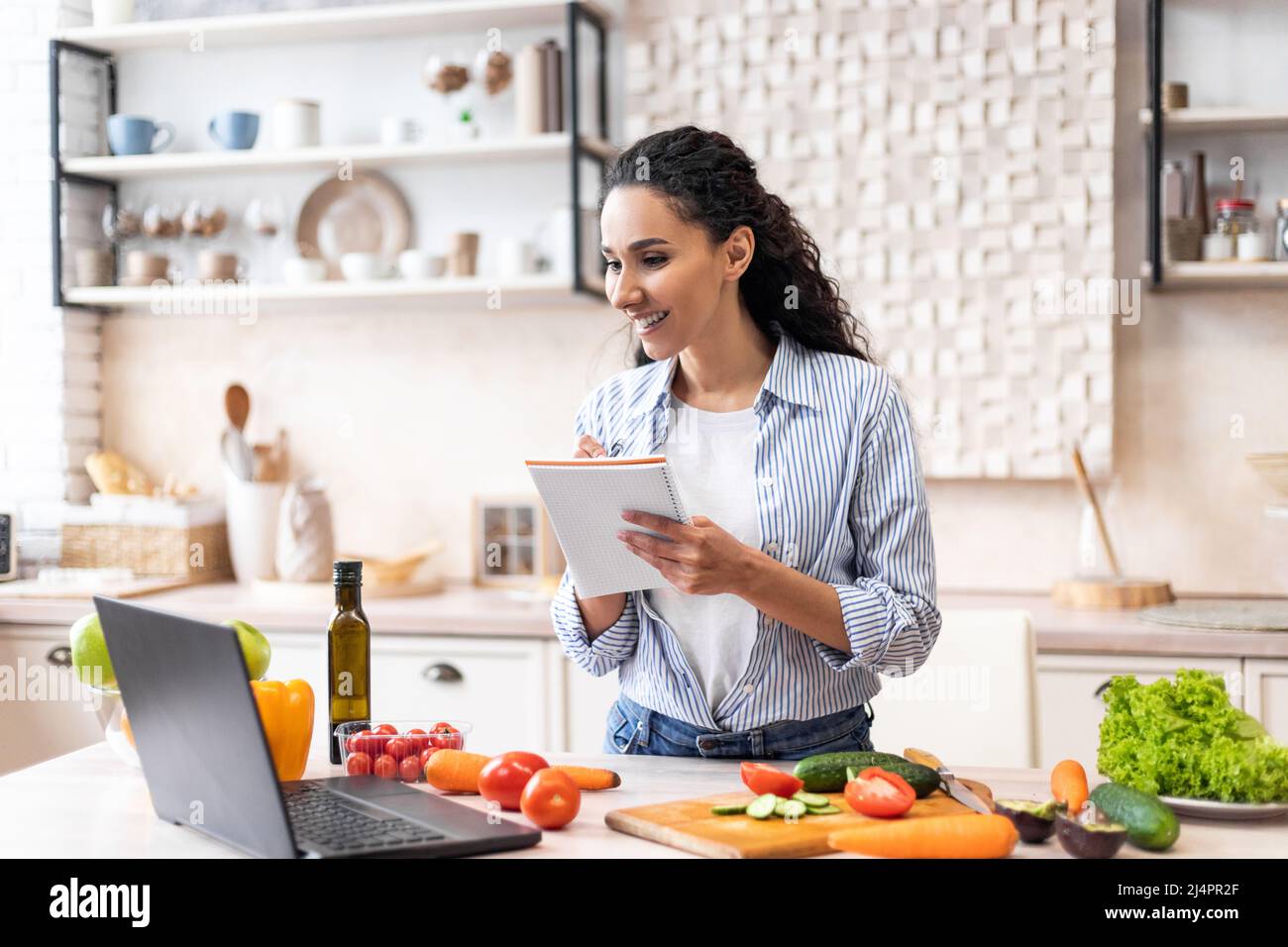 Positive woman writing new recipe while watching video on laptop ...