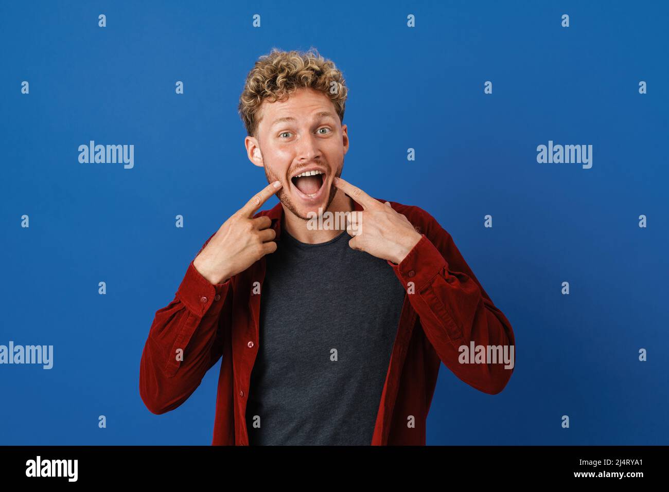 Young blonde man screaming and pointing fingers at his mouth isolated ...