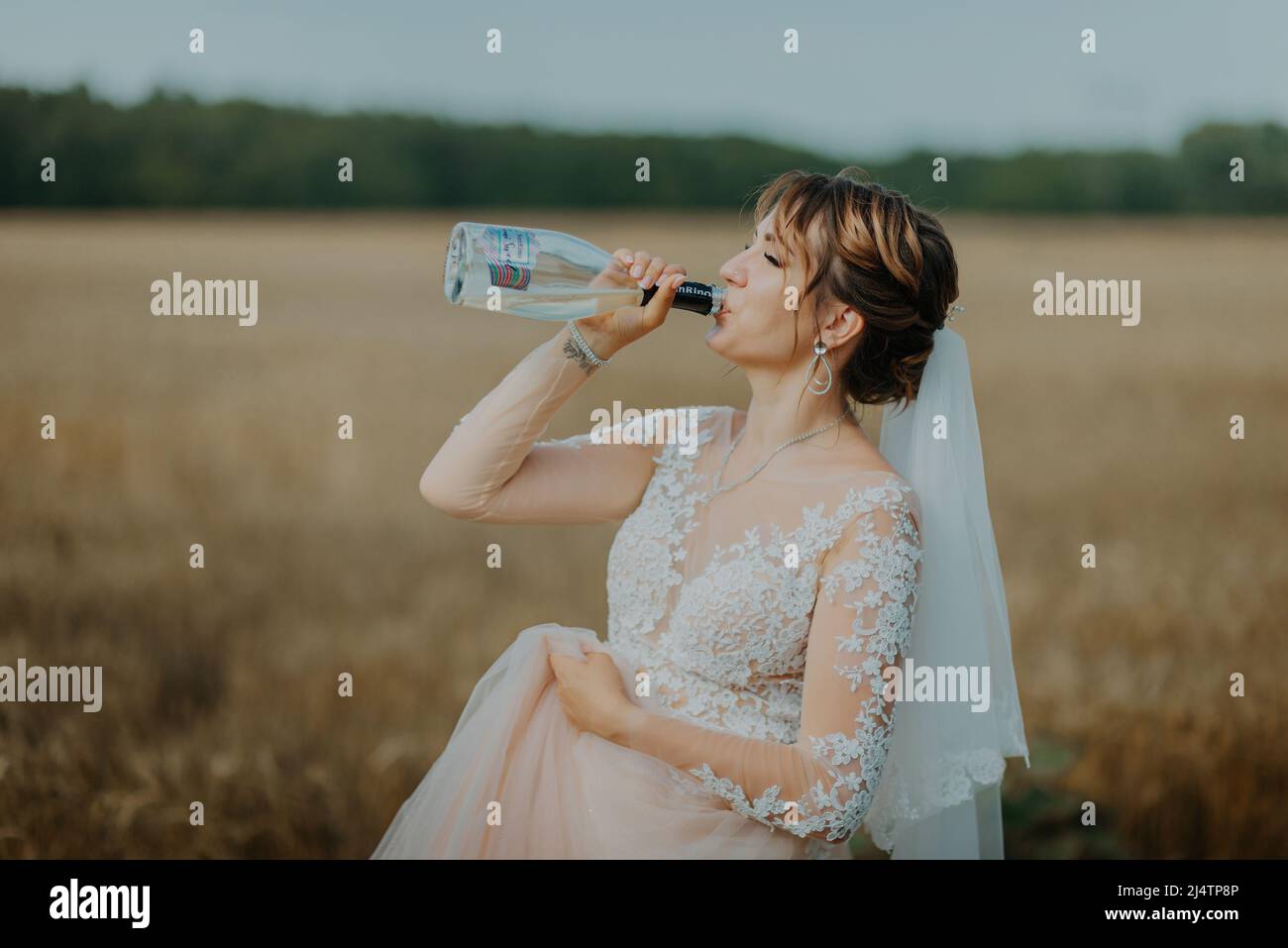 Girl in wedding dress and white veil drinking champagne from bottle ...