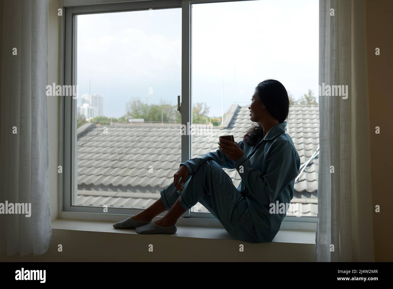 Positive young woman sitting on window sill, drinking morning coffee ...