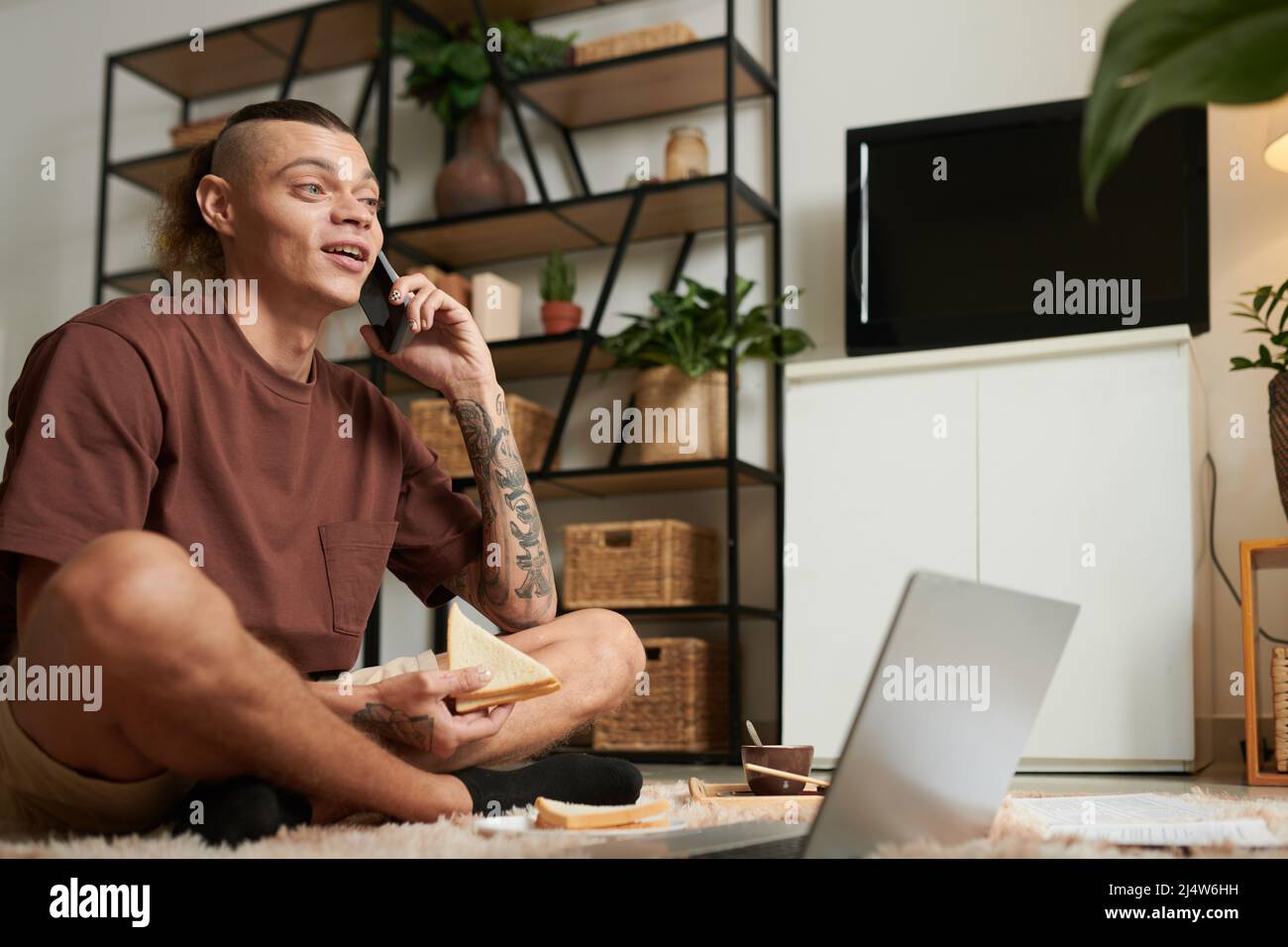 Positive young man eating sandwich and talking on phone when sitting on ...