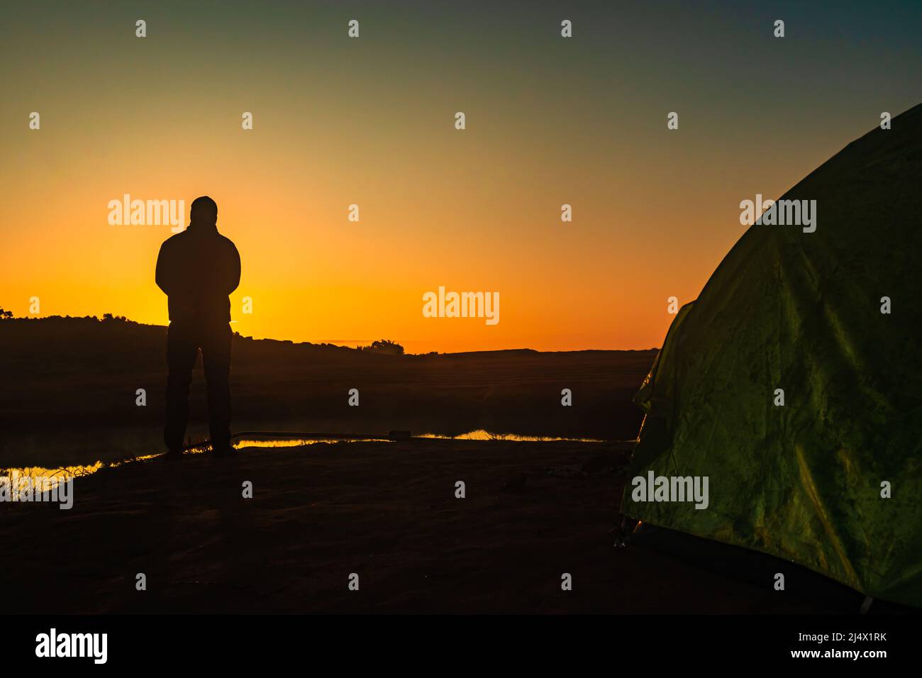 back lit man standing with sunrise over mountain shadow and calm lake ...