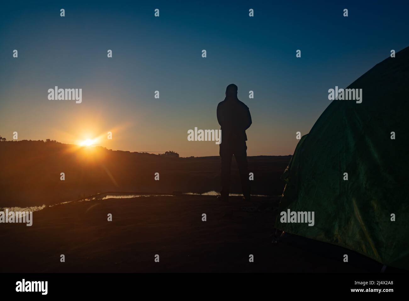 back lit man standing with sunrise over mountain shadow and calm lake ...