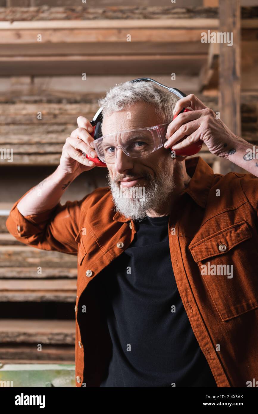 bearded woodworker putting on protective earmuffs and looking at camera ...
