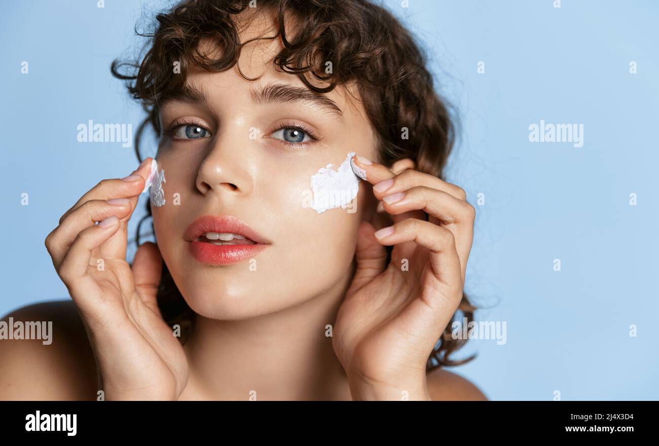 Young woman looking in mirror and applying facial cream on her cheeks ...