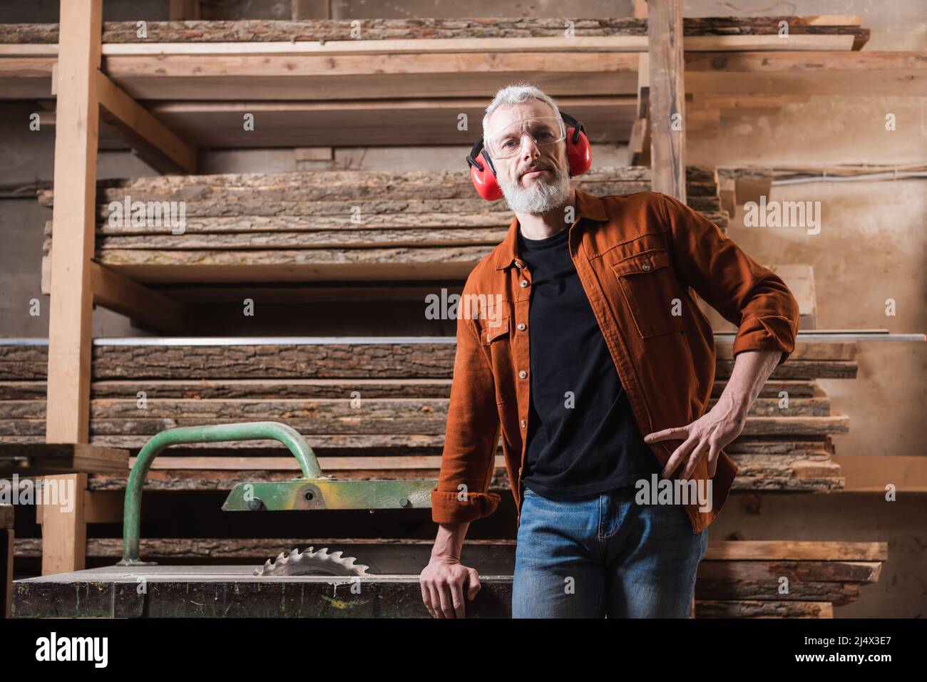 carpenter standing with hand on hip near workbench with circular saw in ...