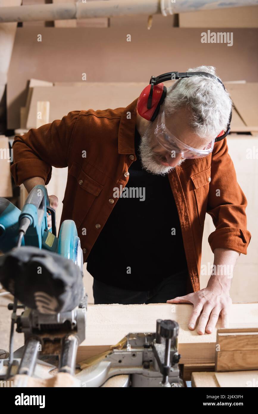 grey haired woodworker cutting timber on miter saw in carpentry studio ...
