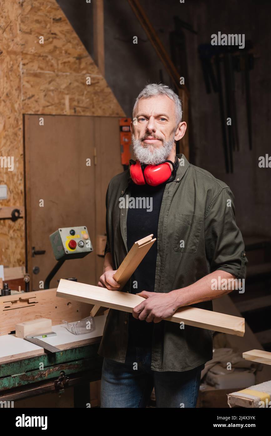 bearded carpenter holding wooden details and looking at camera in ...