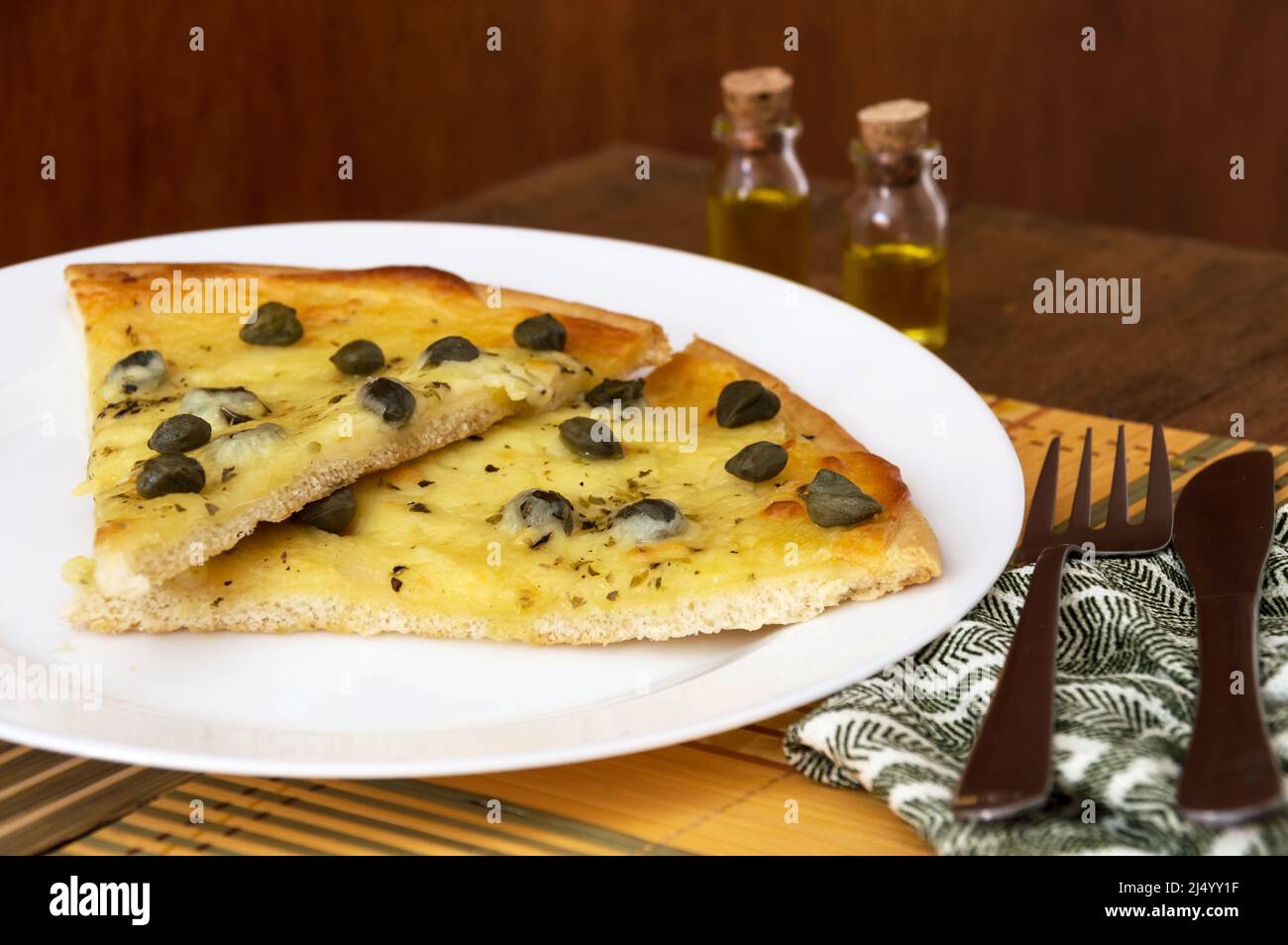 Two slices of caper pizza inside a white plate with cutlery on the side ...