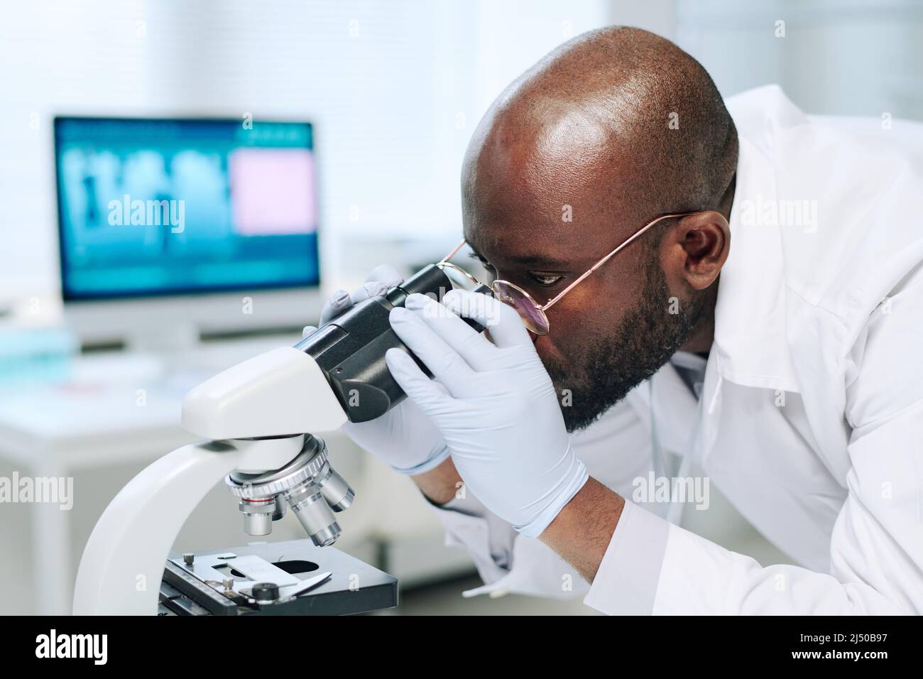 Young serious black man in whitecoat and gloves working with microscope ...