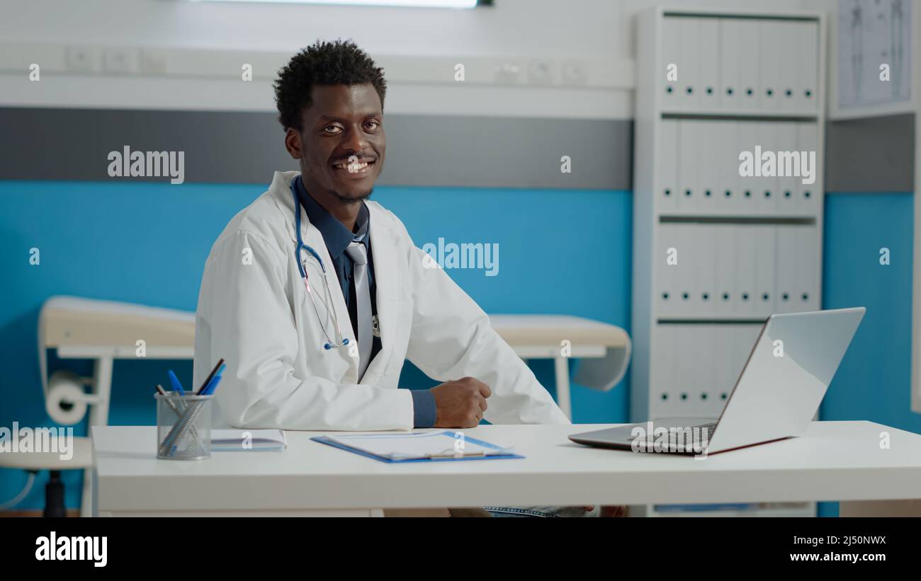 Portrait of african american medic sitting at white desk with laptop ...