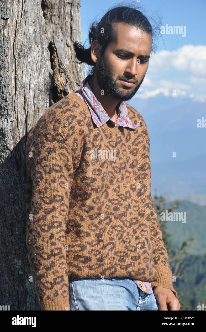 A south asian young man looking down while leaning against tree in the ...