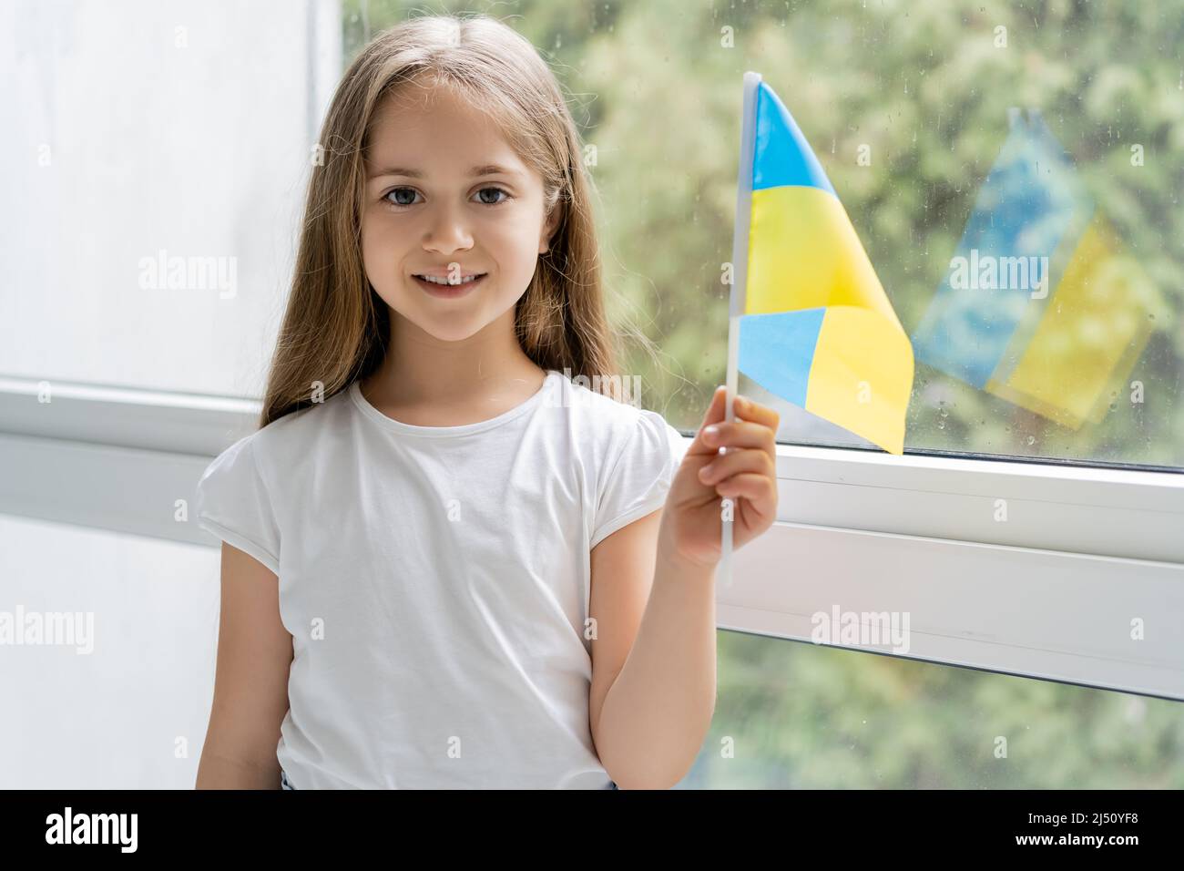 happy girl with small ukrainian flag looking at camera near window at ...