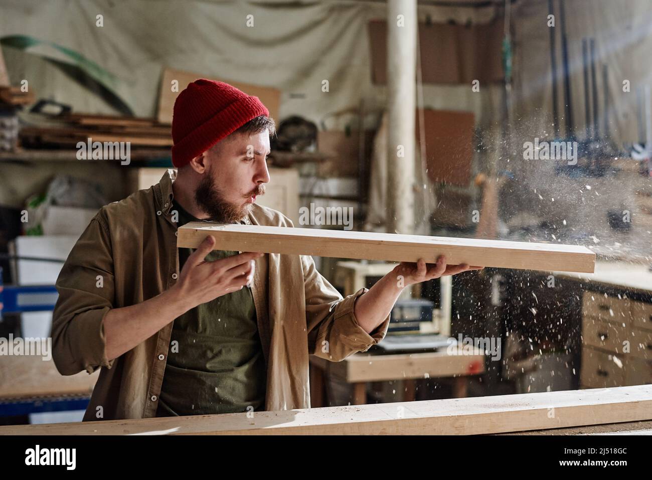 Modern male carpenter wearing red knit cap holding wood plank blowing ...