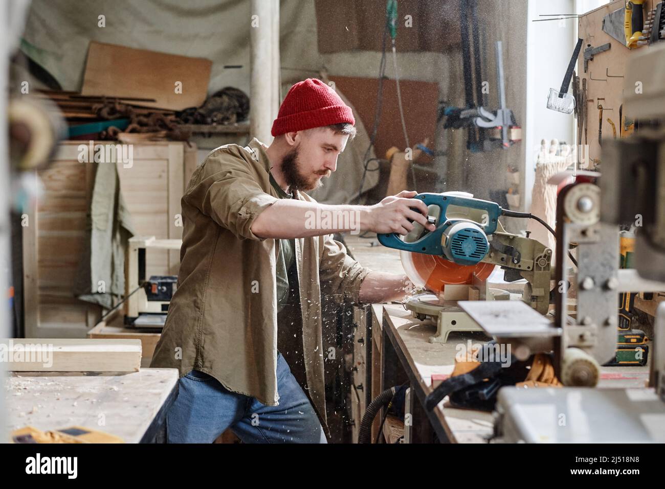 Side view of modern young Caucasian carpenter cutting wooden plank with ...