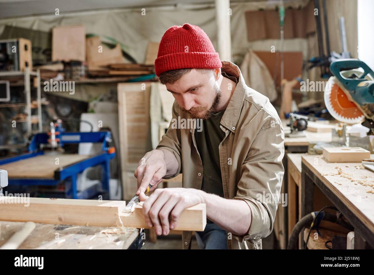 Bearded Caucasian carpenter wearing red knit cap cutting mortise in ...