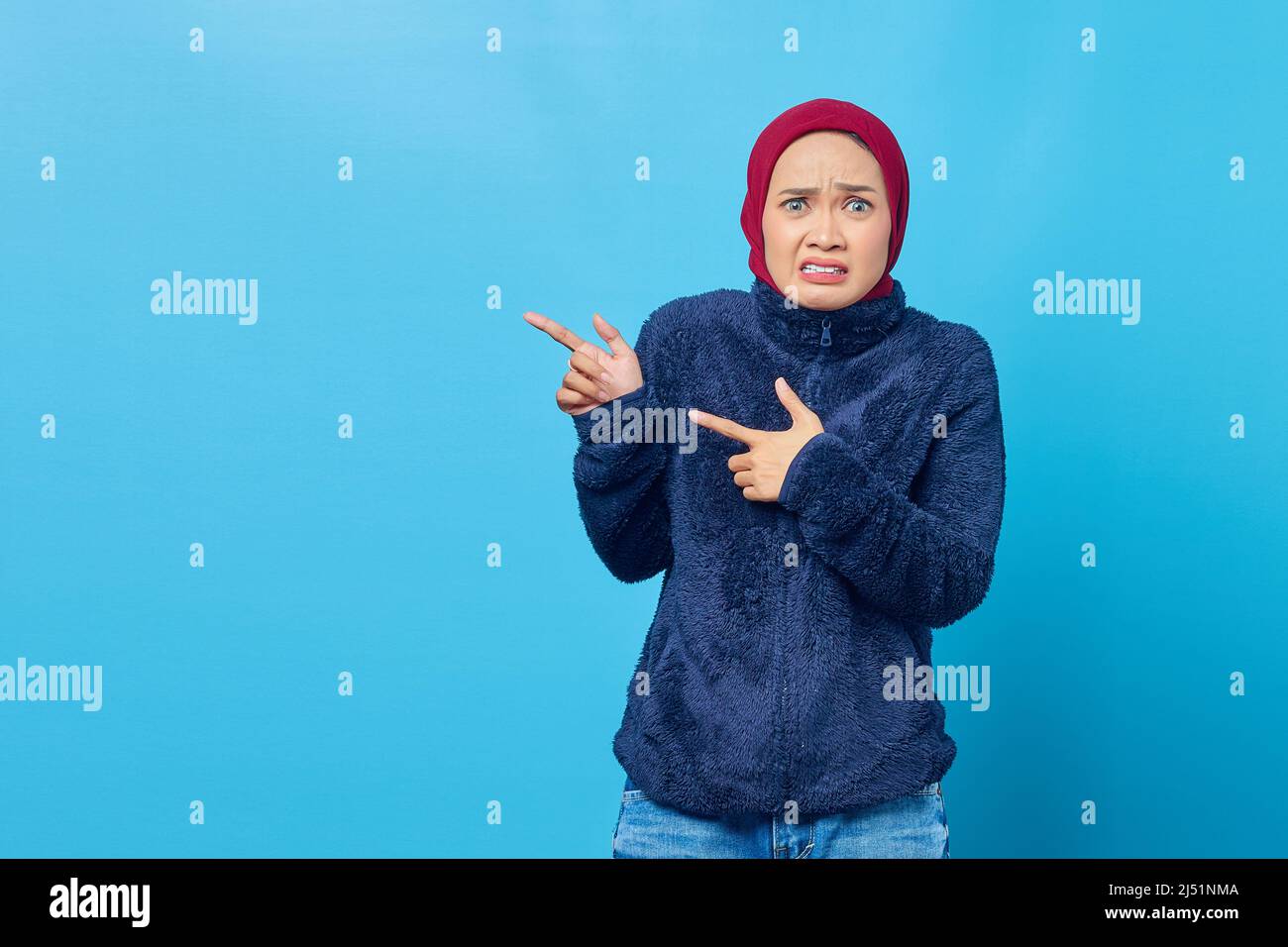 Scared young Asian woman pointing finger at copy space on blue ...
