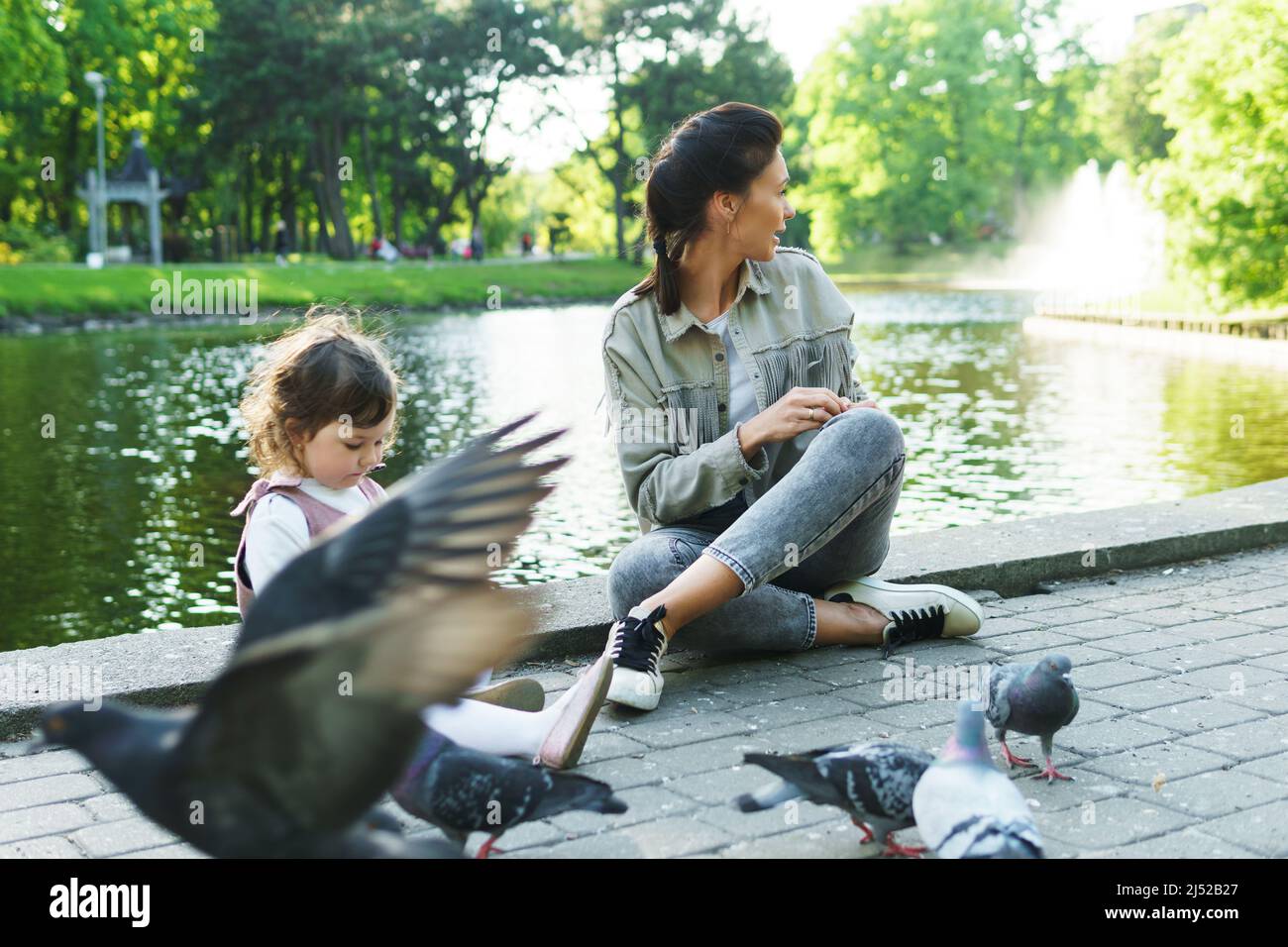 Young mother and her cute little daughter feeding birds in city park ...