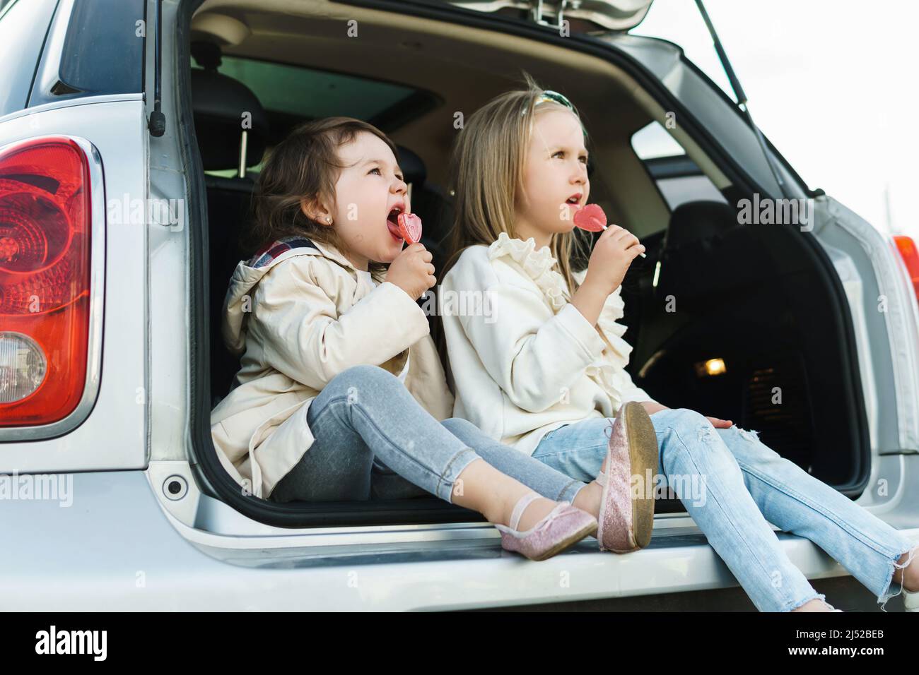 Two little girls sitting in a car's trunk and eating lollipops before a ...