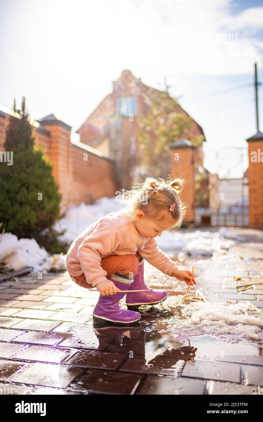 Girl plays in a puddle with wooden stick in spring day at sunlight ...