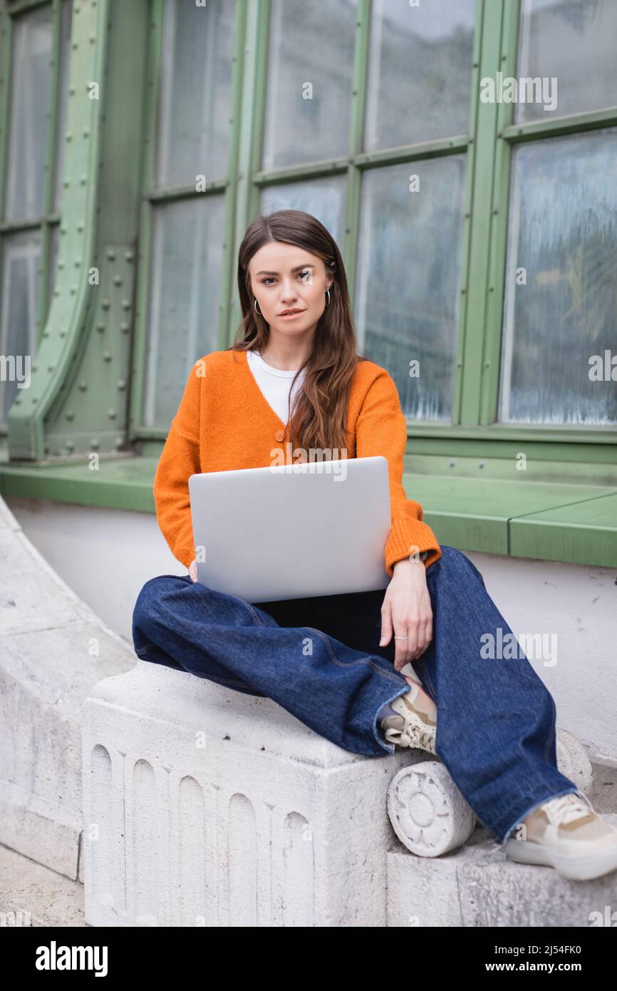 young woman in jeans and orange cardigan sitting with laptop on rooftop ...