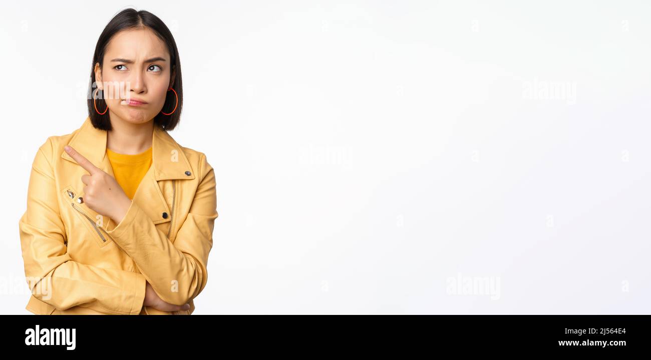 Asian girl looking skeptical, pointing finger left at smth strange, staring doubtful and ...