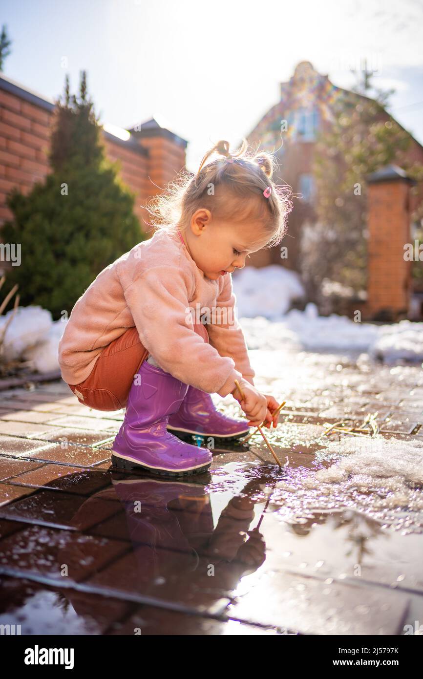 Girl plays in a puddle with wooden stick in spring day at sunlight ...