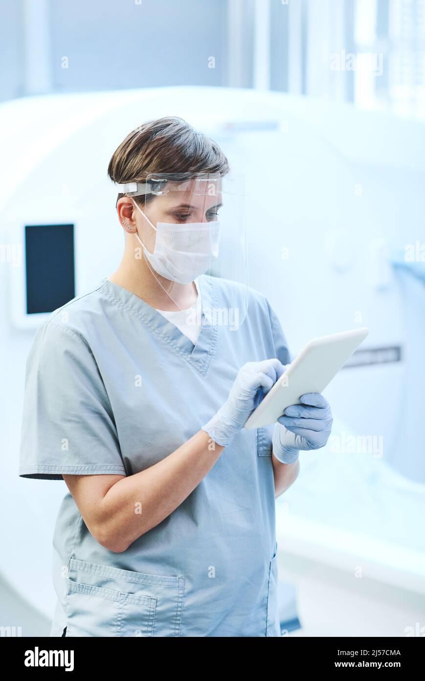 Busy young MRI tech in safety face shield and gloves using tablet while ...