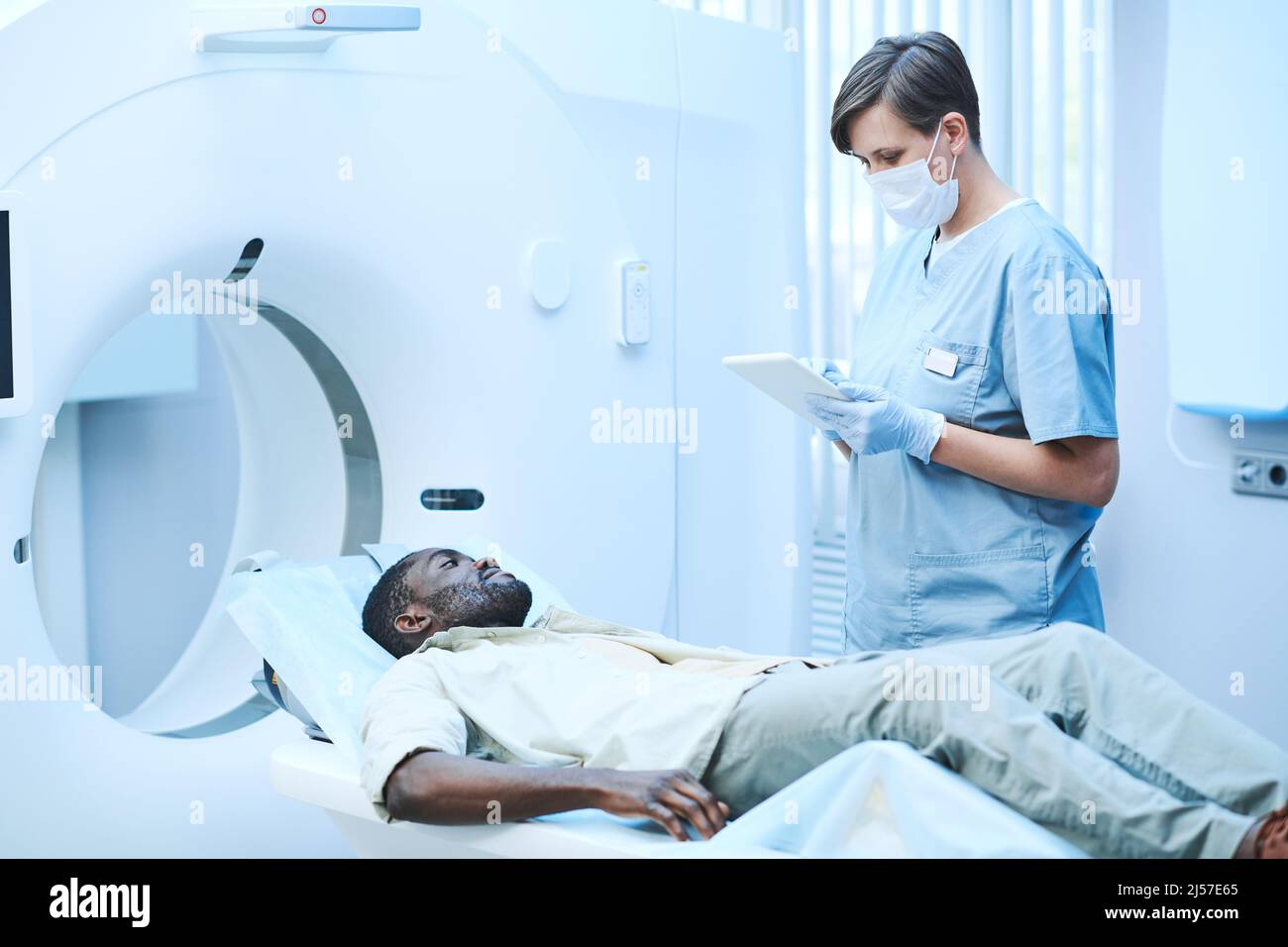 Caucasian nurse in scrubs and mask standing at MRI scanner and asking ...