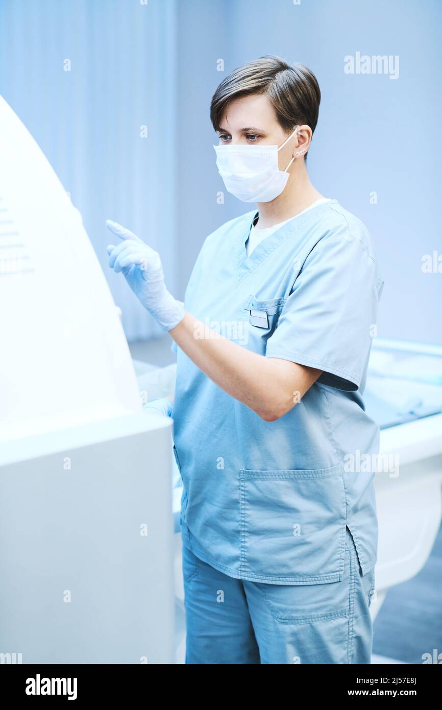 Young MRI technologist in scrubs and mask pushing button on MRI scanner ...