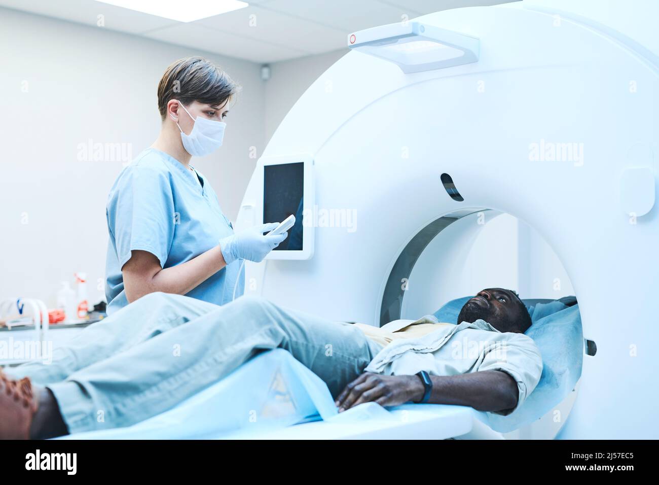 Young Caucasian MRI tech in mask pushing button on control panel while ...