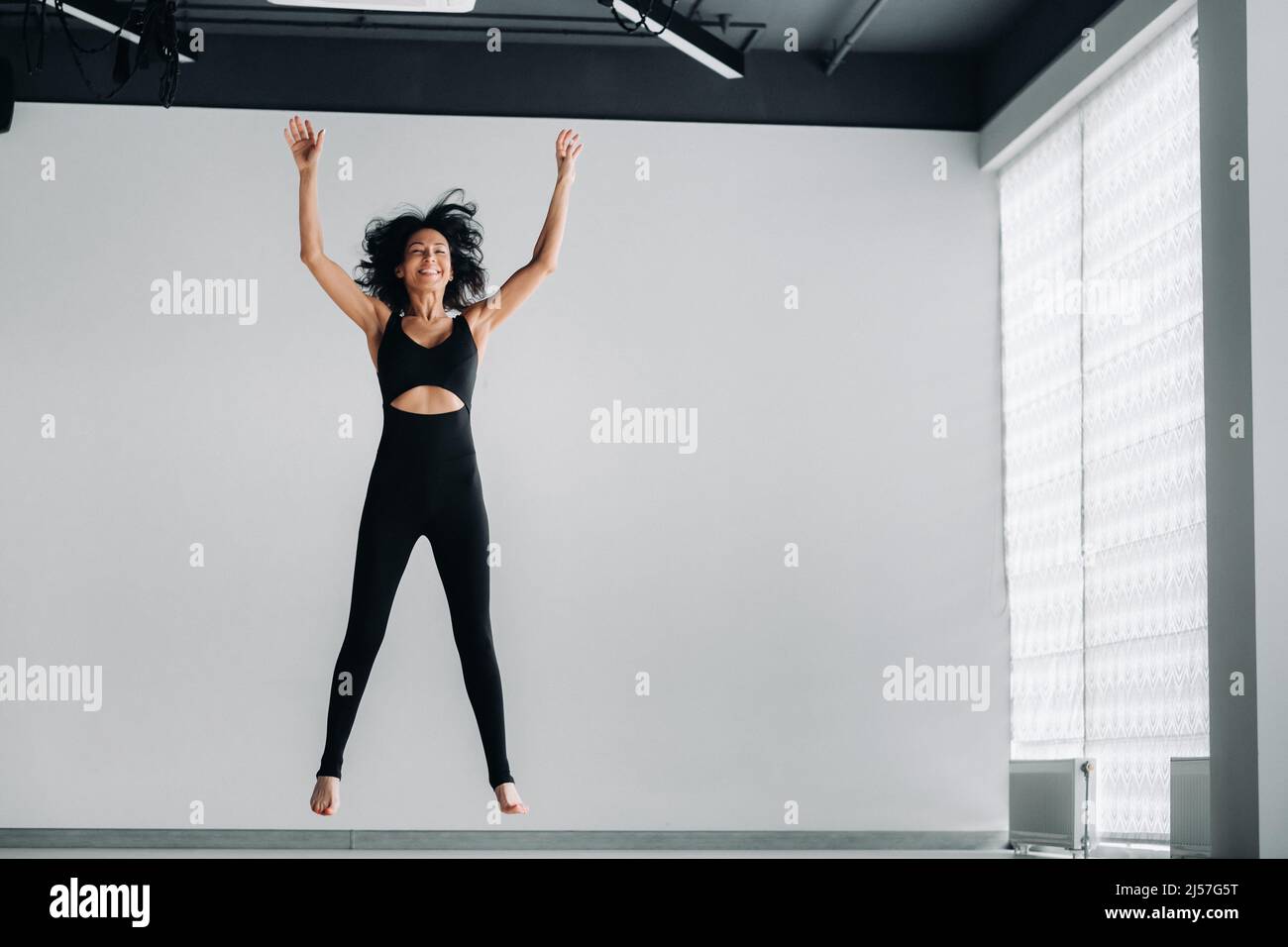 A happy woman in black sportswear makes a jump up on a white background ...