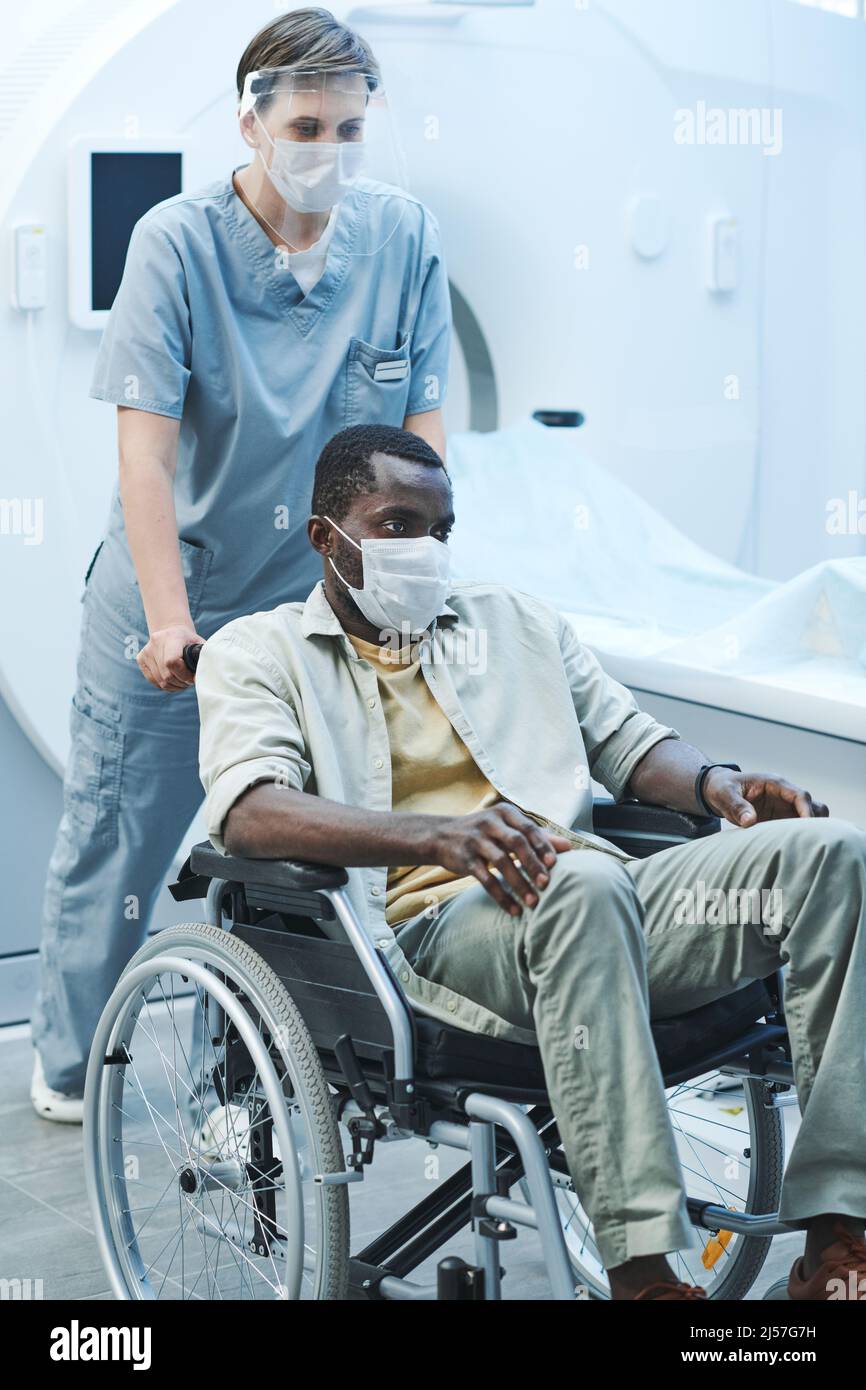 Young nurse in protective face shield pushing wheeled chair with Afro ...