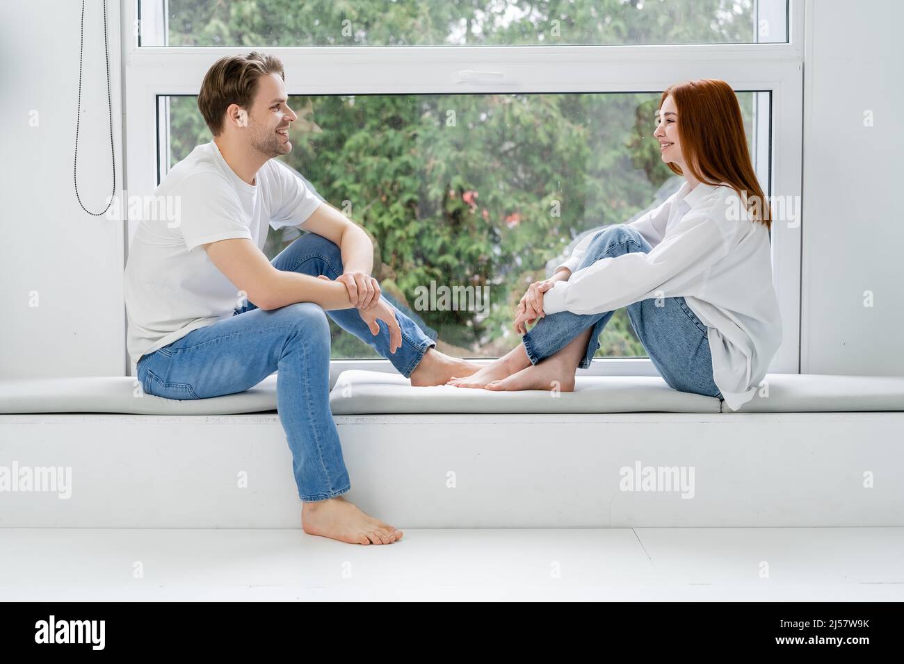 Side view of smiling couple in jeans sitting on windowsill at home ...