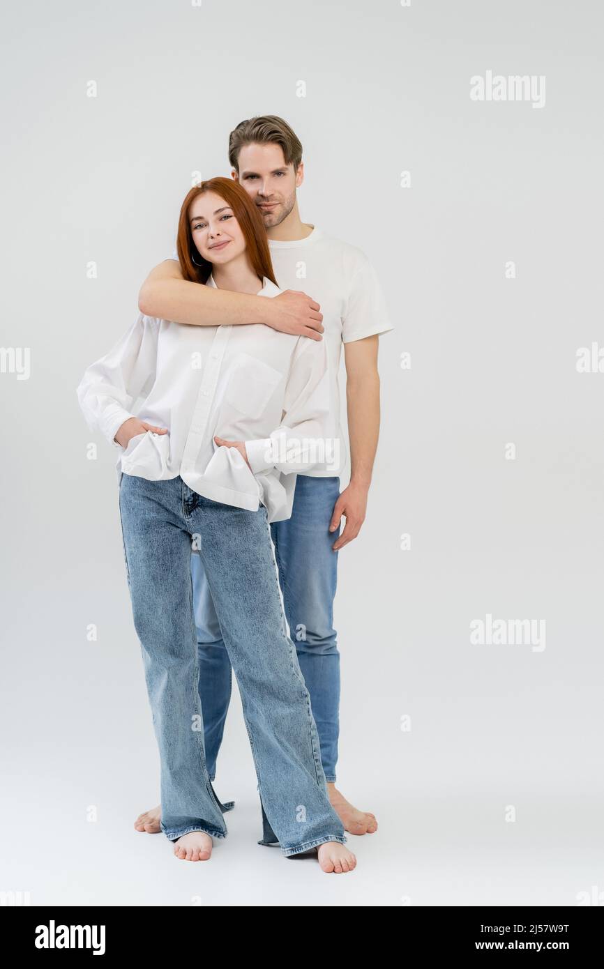 Full length of barefoot man hugging girlfriend and looking at camera on ...