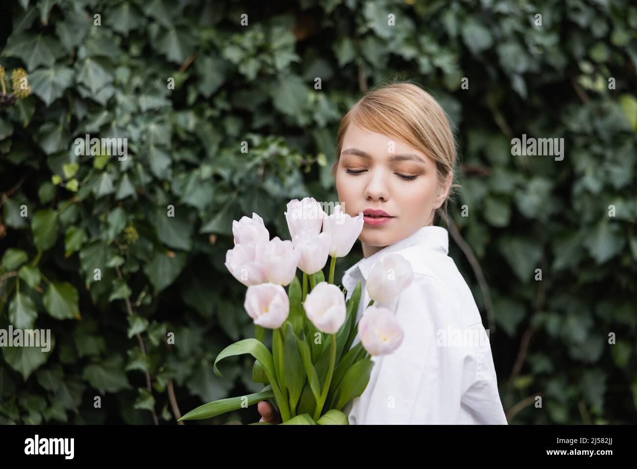 charming woman with bouquet of white tulips standing near green ivy ...